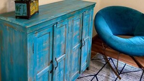 Um armário azul-esverdeado com uma planta, uma cadeira azul e um espelho colorido na parede.