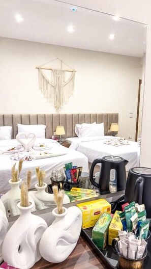 Quarto de hotel com duas camas, um painel de macramé na parede e uma bandeja com produtos de higiene e alimentos.
