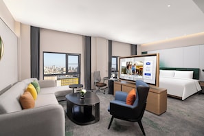 Quarto de hotel com sofá, poltrona, mesa de centro, televisão, cama e vista para a cidade.