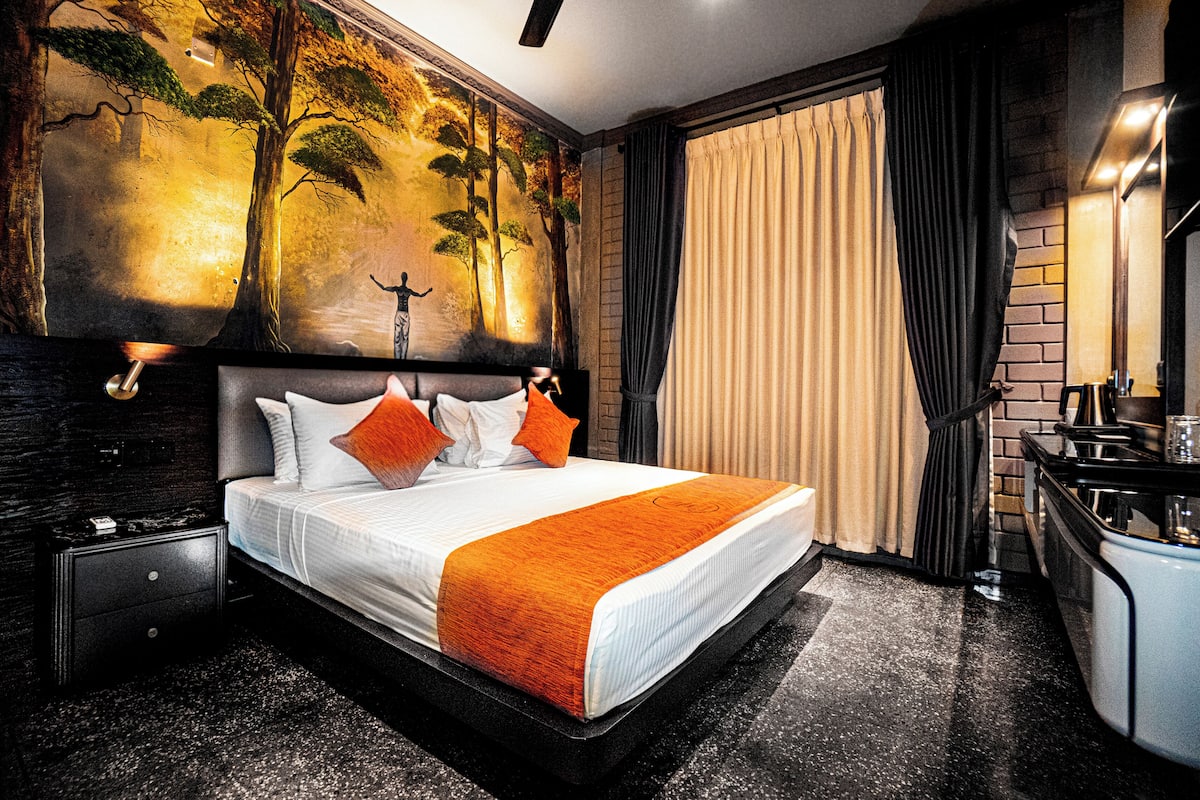 Quarto de hotel com cama, mesa de cabeceira, escrivaninha com cadeira, um quadro na parede e janela com cortinas.