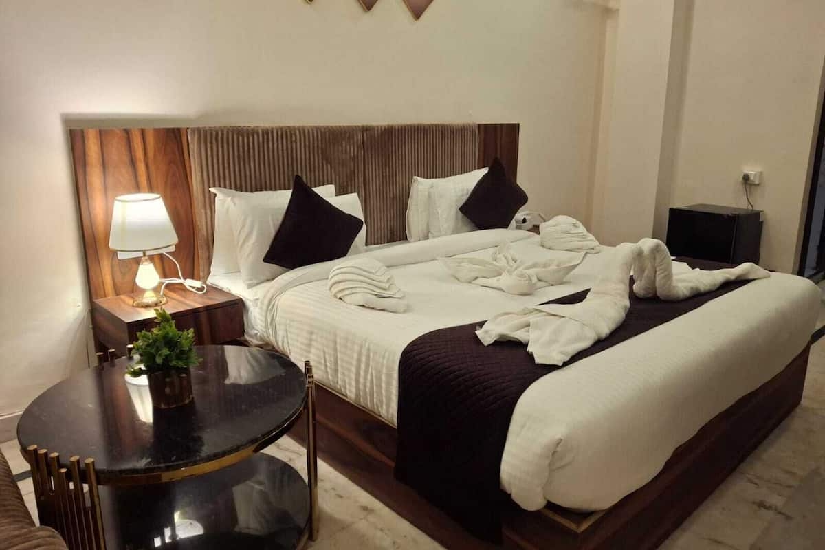 Quarto de hotel com cama, mesa de cabeceira com abajur, mesa redonda com planta e um quadro na parede.