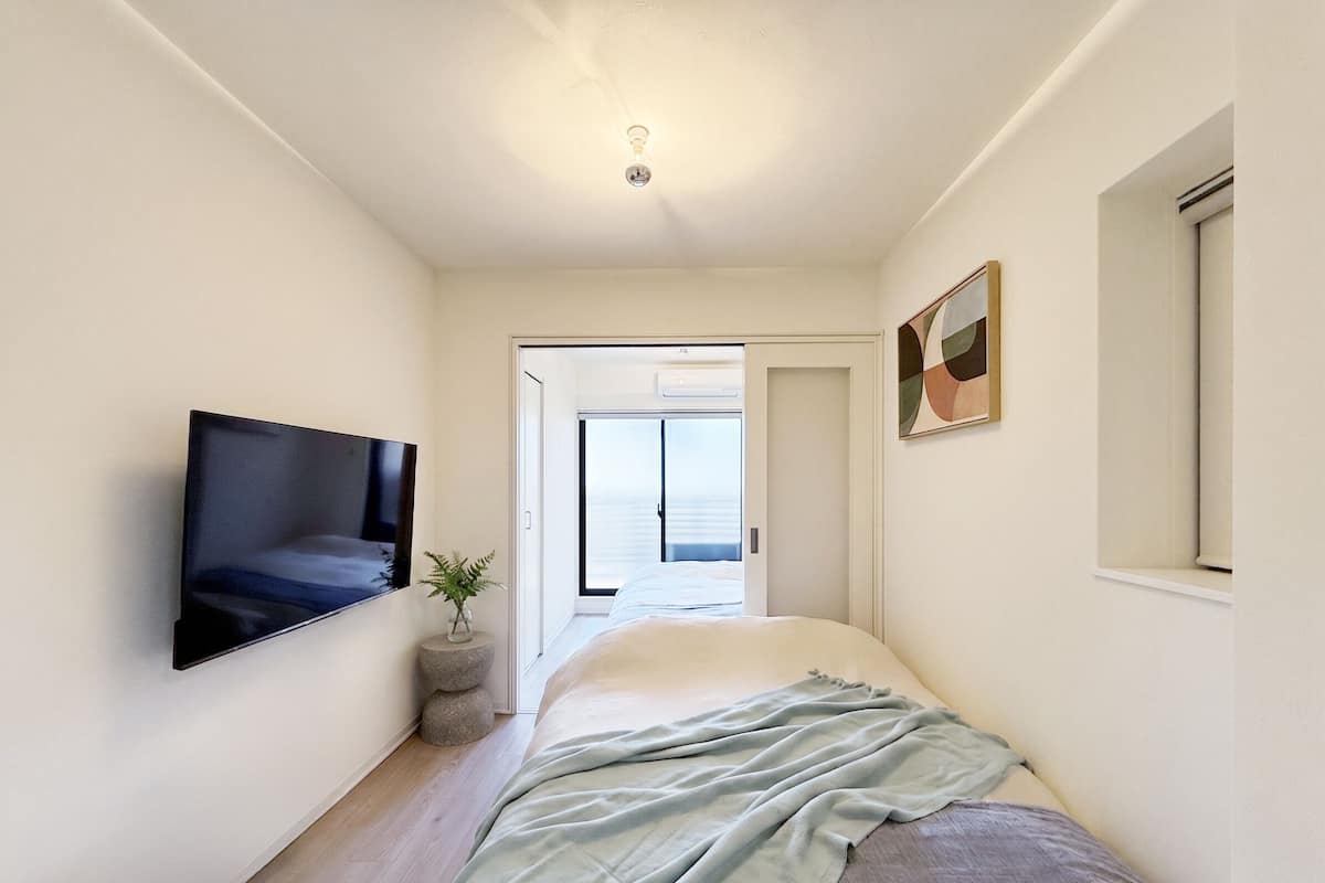Um corredor estreito que leva a um quarto com uma cama, uma televisão na parede, um vaso de planta e um quadro.