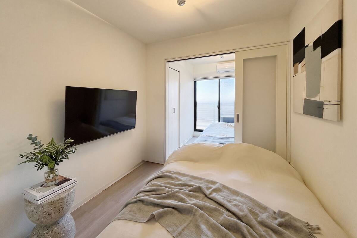 Quarto com cama, televisão de tela plana, planta em vaso e vista através de uma porta aberta.