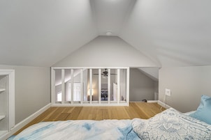 Quarto com teto inclinado, piso de madeira, cama com roupa de cama estampada e vista para um cômodo adjacente através de uma divisória branca.