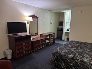 Quarto de hotel com cama, escrivaninha, televisão, cômoda, espelho e cozinha compacta.