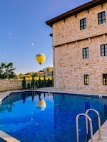 Um edifício de pedra com piscina, balões de ar quente ao fundo e céu claro.