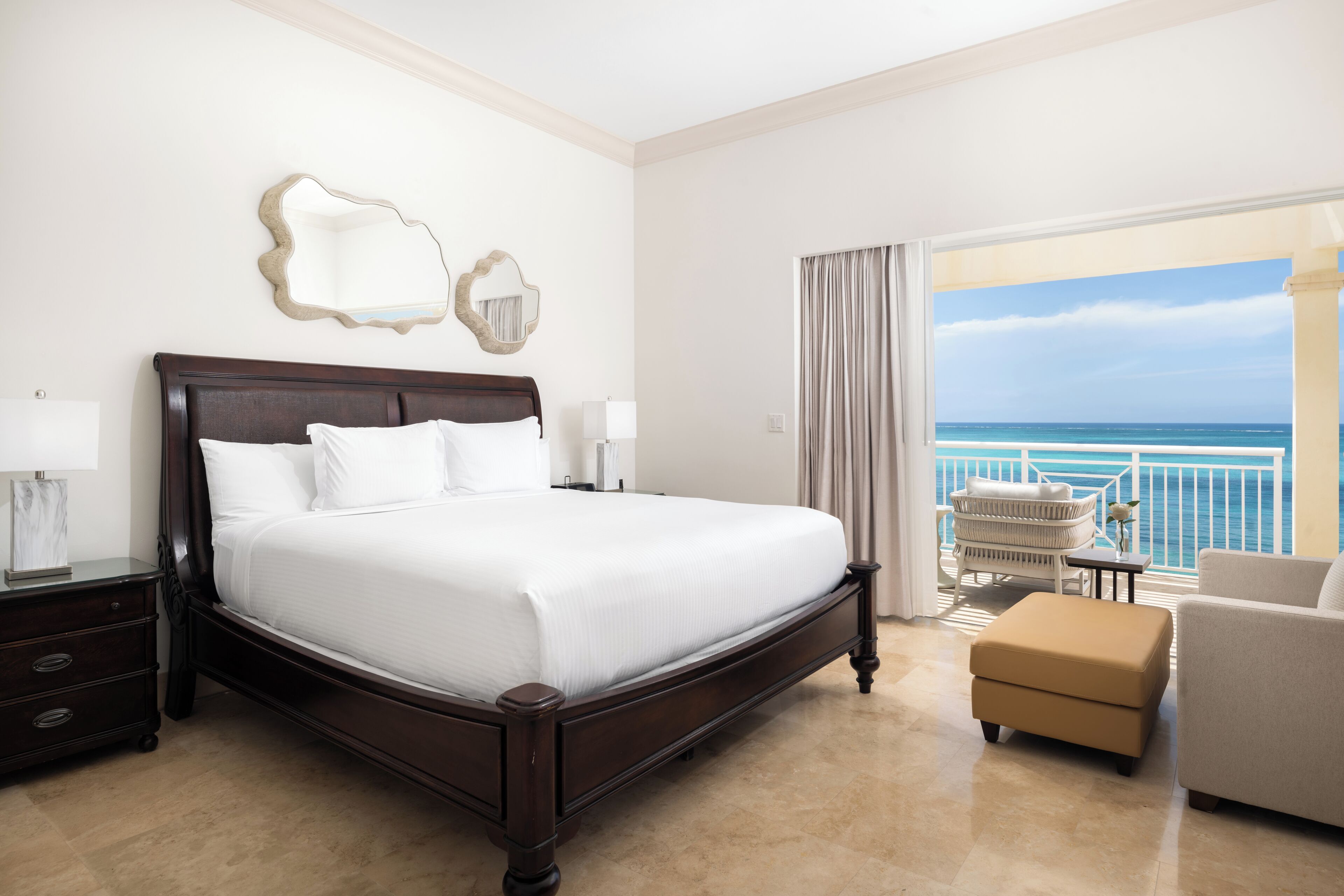 Quarto de hotel com uma cama grande, duas mesas de cabeceira, uma cadeira, uma mesinha e varanda com vista para o mar.