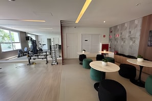Uma academia com equipamentos de ginástica, uma área de refeições com mesas e cadeiras, e uma grande janela que ilumina naturalmente o espaço.