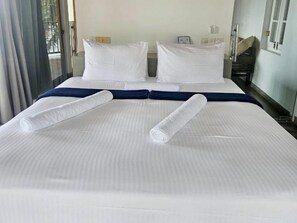 Cama bem arrumada com lençóis brancos e duas toalhas enroladas, mesa de cabeceira com abajur e janela com cortinas.