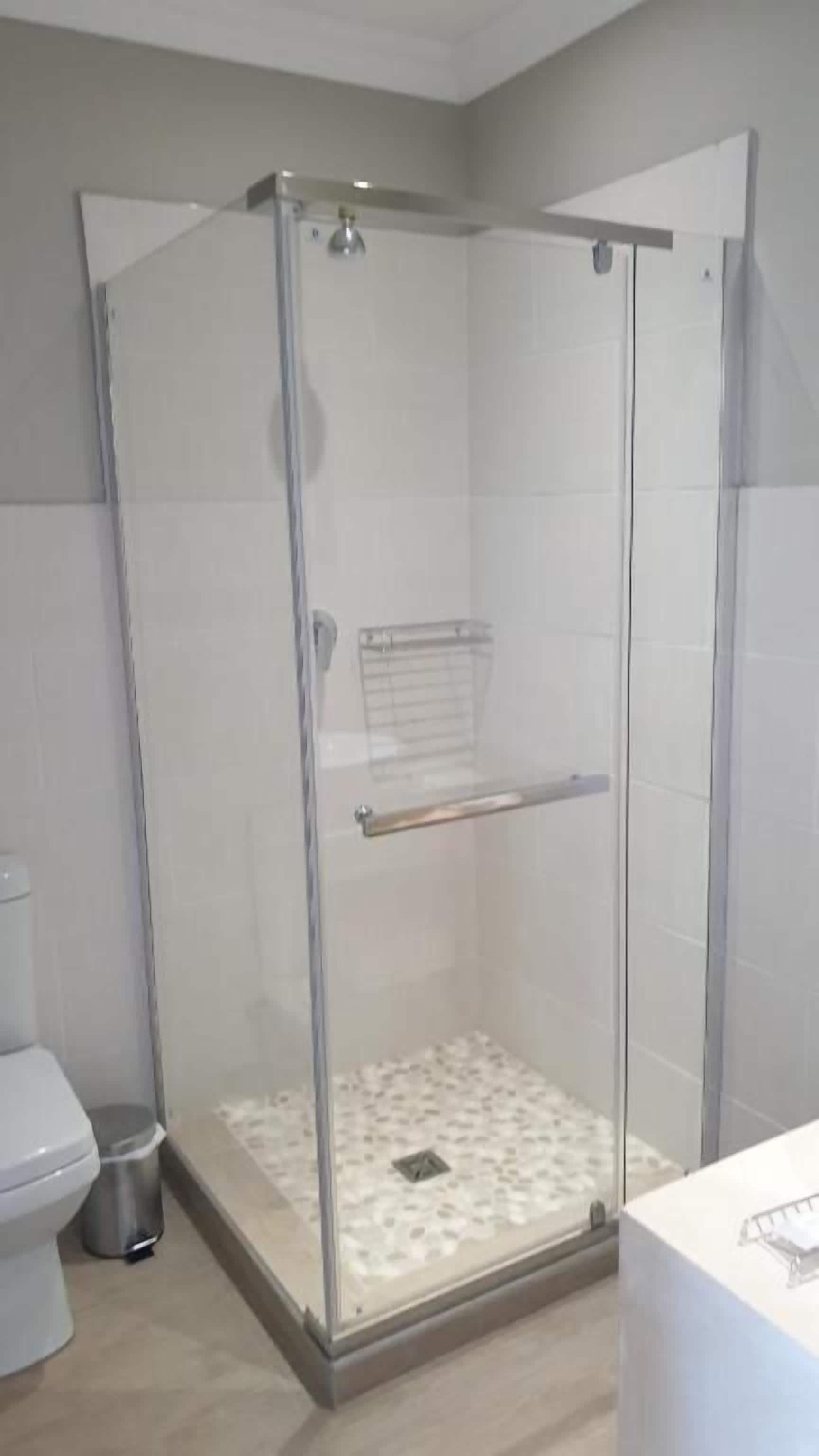 Um chuveiro com paredes de vidro, uma prateleira de metal, vaso sanitário e lixeira em um banheiro com paredes e piso revestidos de azulejo.