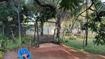 Um baloiço, um banco e um caminho de terra num jardim com árvores e plantas em vasos.