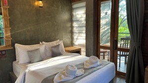 Quarto de hotel com cama, almofadas, mesa de cabeceira, janela com persianas e vista para o exterior com vegetação.