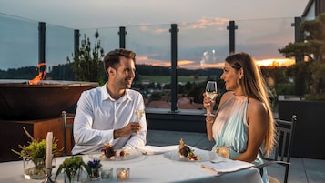 Um casal jantando ao ar livre com vista para um pôr do sol e uma lareira ao fundo.