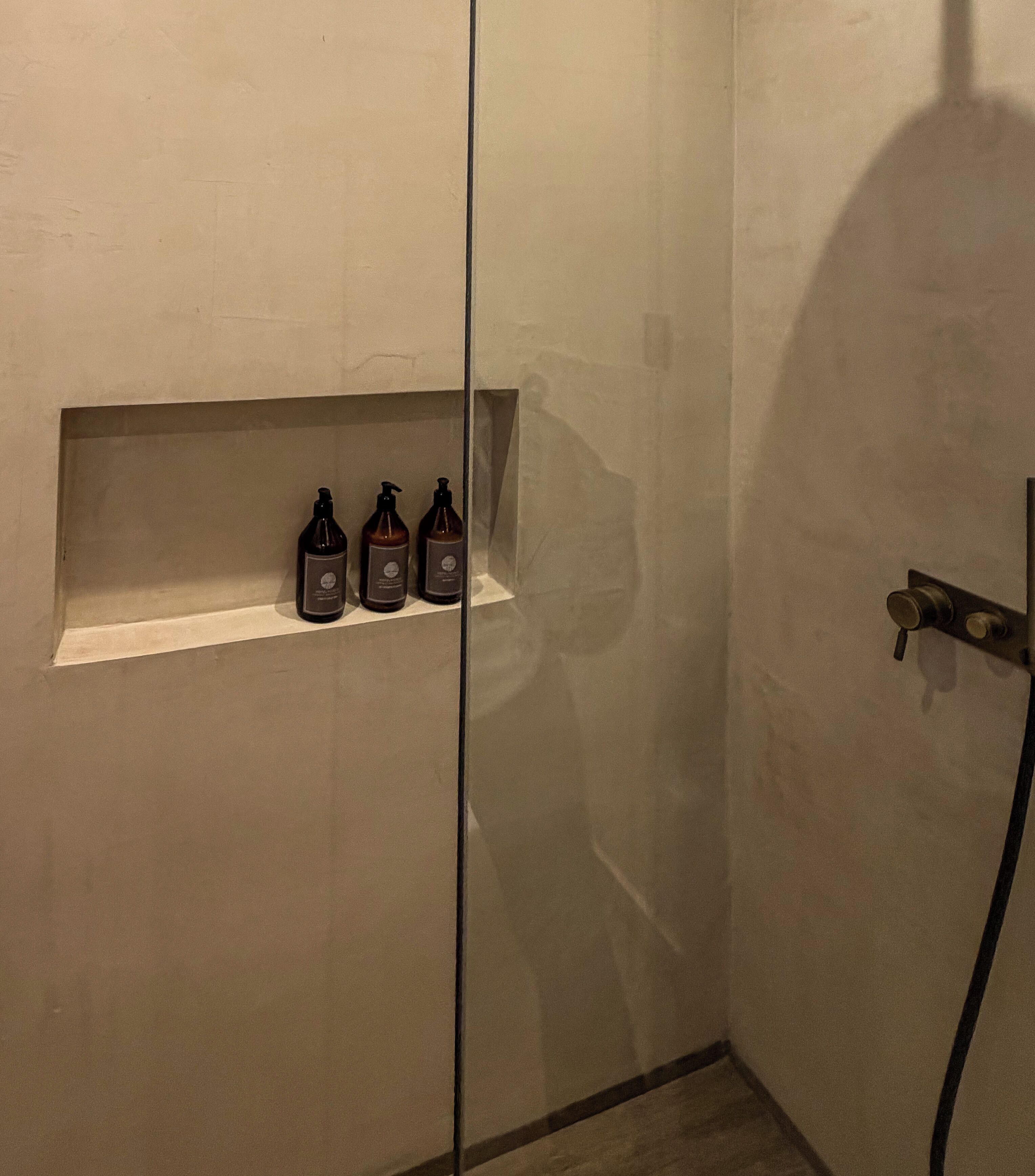 Casa de banho com divisória de vidro, uma prateleira com três garrafas e porta de chuveiro.