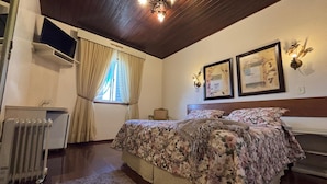 Quarto com teto de madeira, cama com roupa de cama florida, radiador e duas luminárias fixadas na parede.