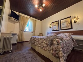 Quarto com teto de madeira, cama com roupa de cama florida, radiador e duas luminárias fixadas na parede.