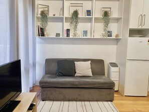 Uma sala de estar moderna com um sofá cinzento, uma televisão de tela plana, um frigorífico branco e uma estante com objetos decorativos.