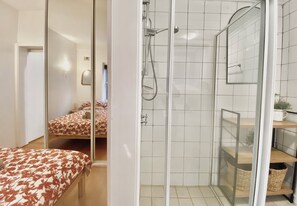 Quarto com cama, chuveiro em box de vidro e pequena cozinha com pia e prateleiras.