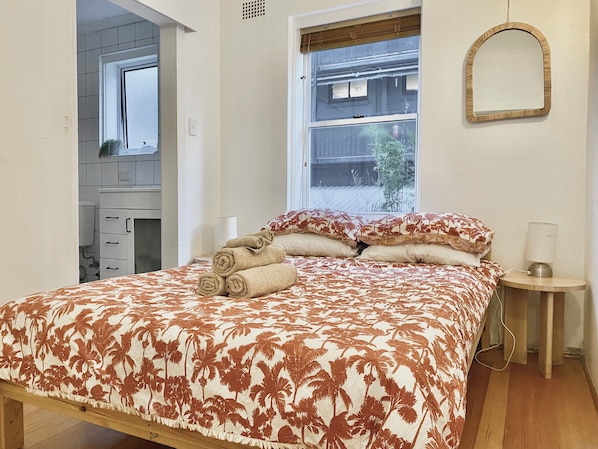Quarto com cama, mesa de cabeceira, abajur, janela com vista e banheiro visível por uma porta aberta.