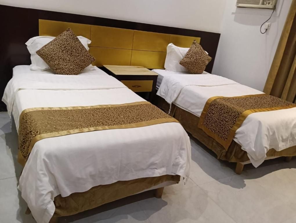 Quarto de hotel com duas camas, cada uma com colcha branca e manta estampada marrom. Há uma mesa de cabeceira de madeira entre as camas, com uma luminária.