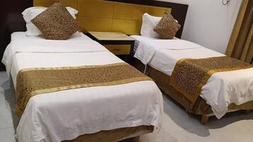 Quarto de hotel com duas camas, cada uma com colcha branca e manta estampada marrom. Há uma mesa de cabeceira de madeira entre as camas, com uma luminária.