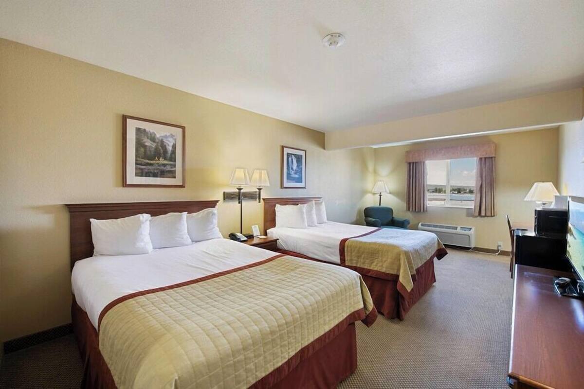 Quarto de hotel com duas camas, uma escrivaninha, uma cadeira, janela com cortinas e duas luminárias na parede.
