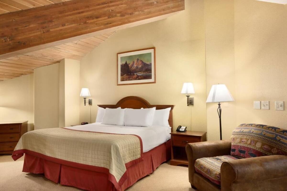 Quarto de hotel com teto de madeira, cama com roupa de cama branca, duas luminárias de cabeceira, uma poltrona estampada e uma pintura de paisagem montanhosa na parede.