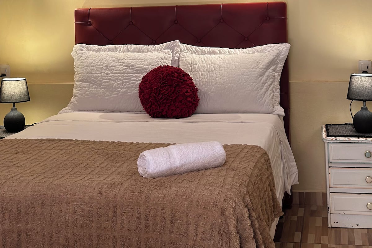 Cama bem arrumada com um travesseiro vermelho, um travesseiro branco e um cobertor marrom. Há uma moldura com uma imagem de um círculo vermelho na parede acima da cama.