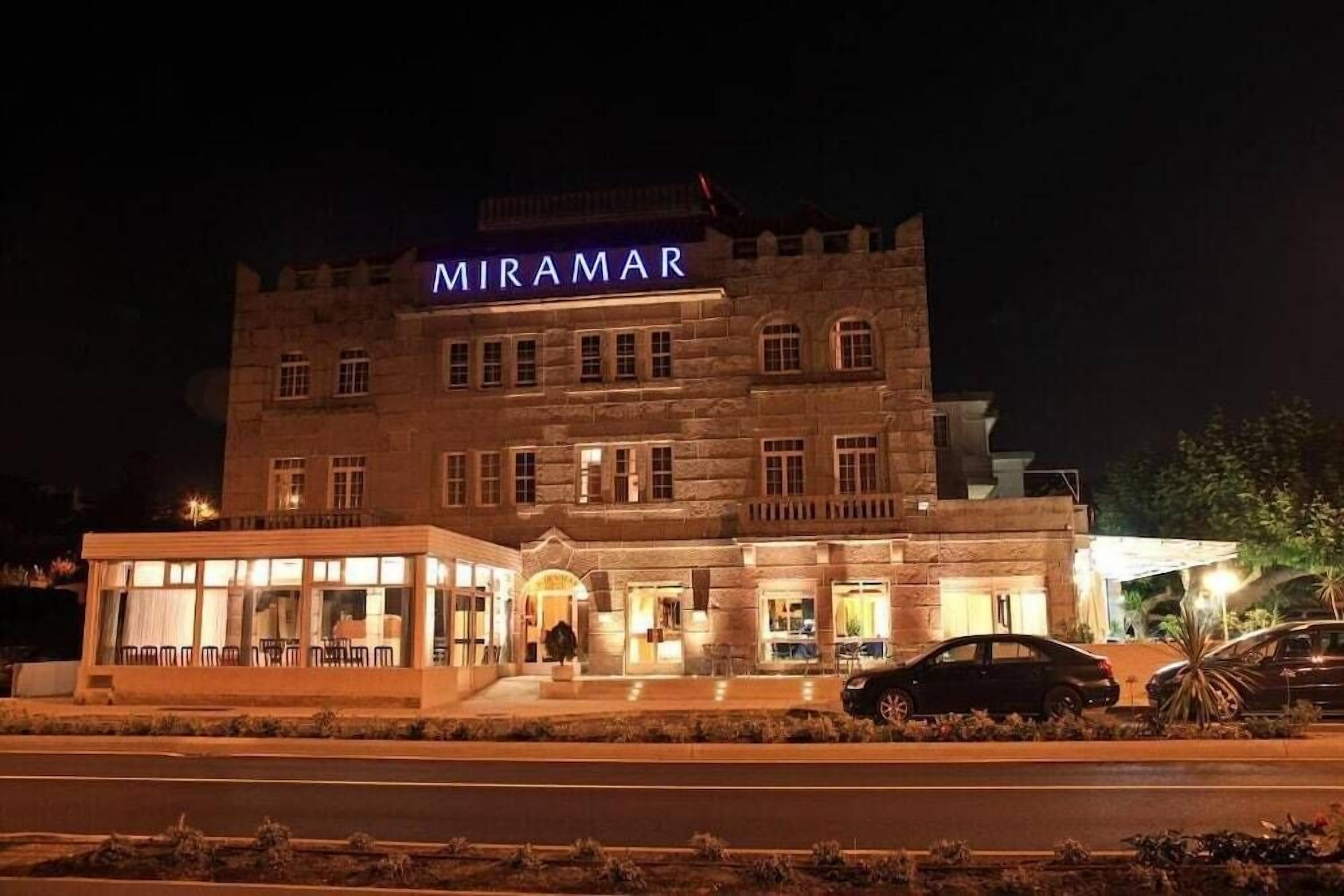 Um hotel chamado MIRAMAR com fachada de pedra, entrada de vidro moderna e área de estacionamento com carros.