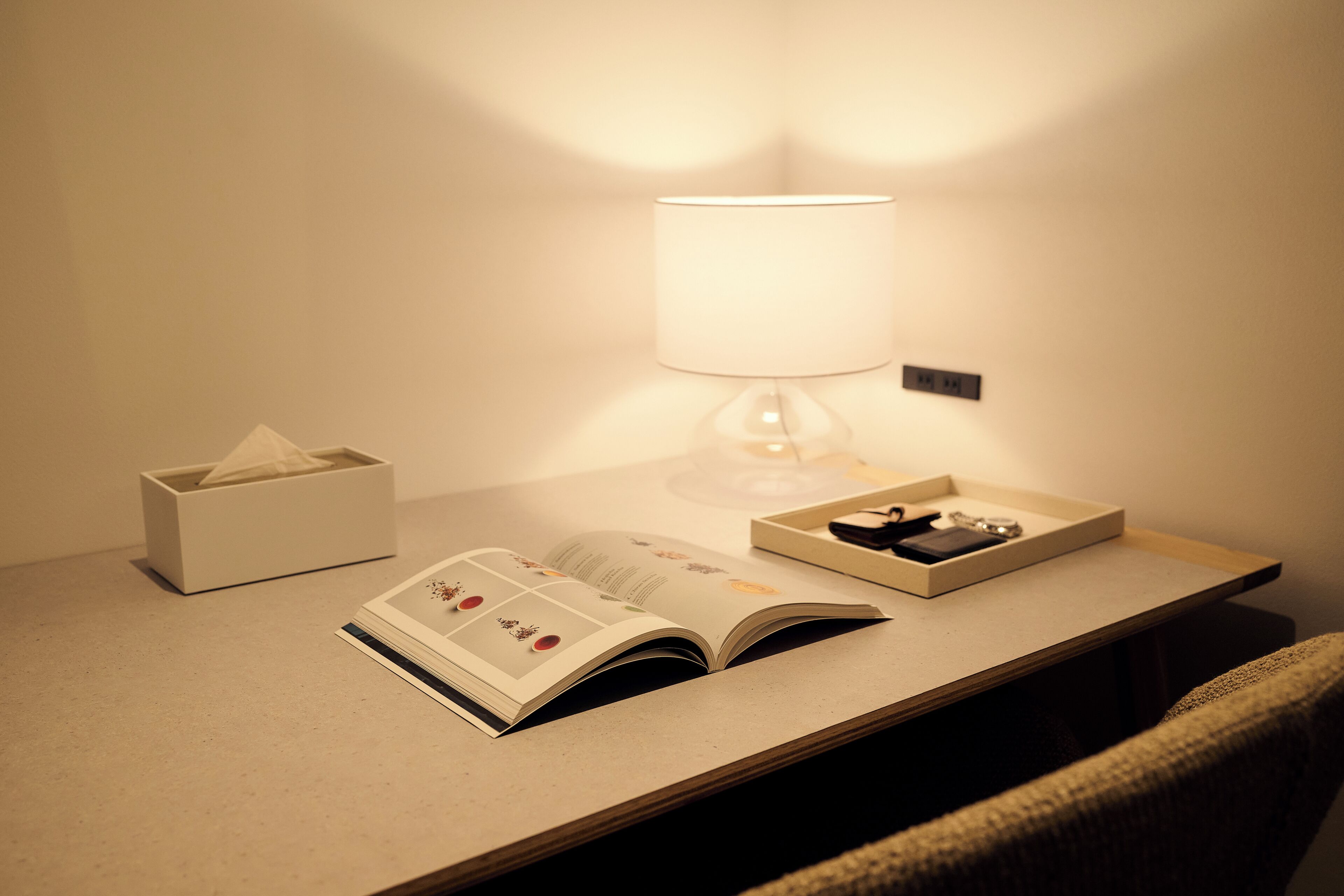 Quarto de hotel bem arrumado com uma mesa de cabeceira, uma luminária, um estojo de lenços de papel, um livro e uma bandeja com uma câmera e um par de óculos.