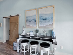 Uma área de bar com balcão de madeira, três banquetas e duas pinturas emolduradas com tema de praia.