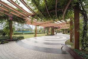 Uma galeria coberta com bancos, rodeada por vegetação e um pavimento ornamental.
