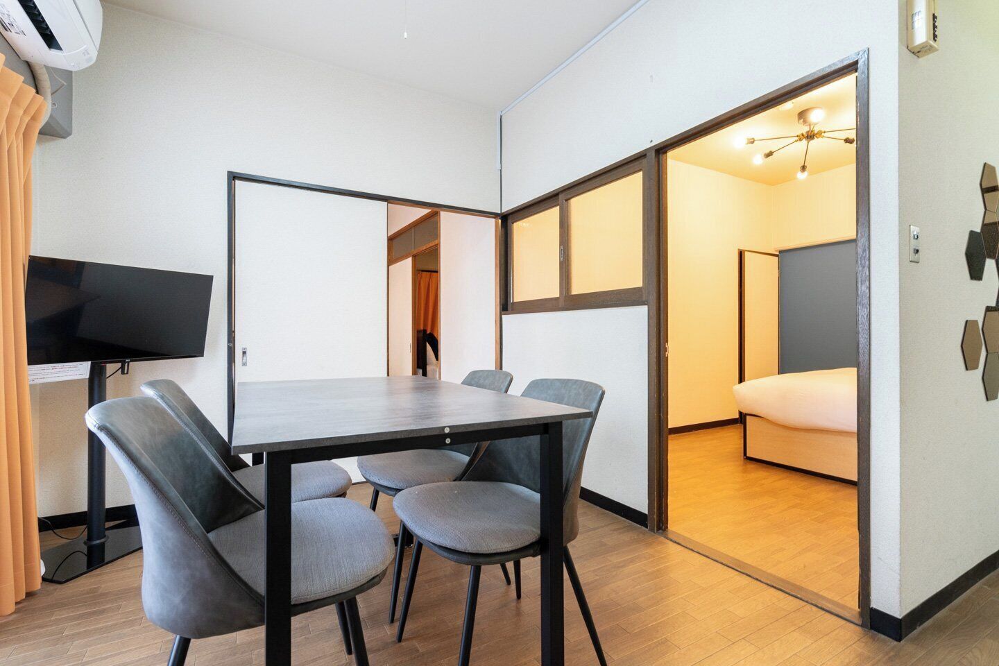 Quarto moderno com televisão de tela plana, mesa de jantar com cadeiras, uma cama e porta deslizante para outro cômodo.