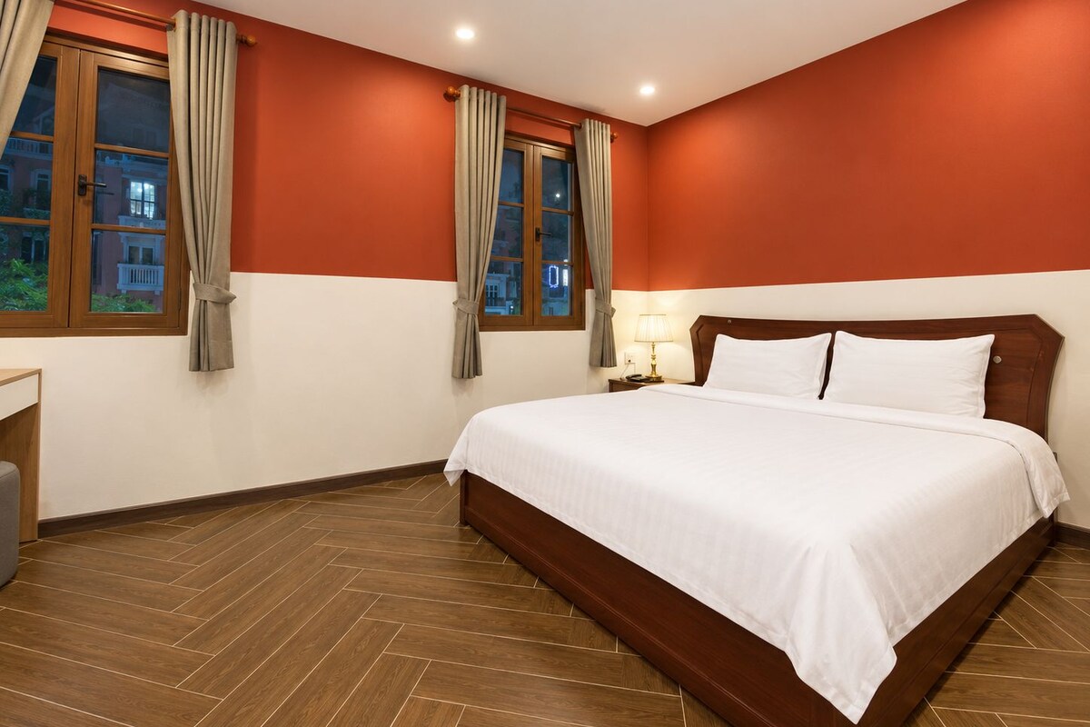 Quarto de hotel com uma cama grande, duas janelas com cortinas, uma mesa de cabeceira com abajur e vista para prédios.