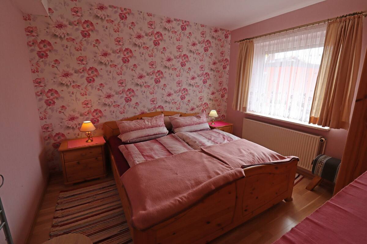 Quarto com cama de madeira, mesa de cabeceira com abajur, radiador e janela com cortinas.