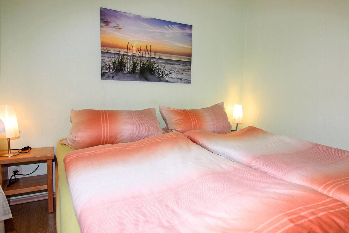 Uma cama bem arrumada com roupa de cama rosa, duas luminárias de cabeceira, uma mesa de cabeceira com abajur e uma pintura de uma paisagem praiana na parede.