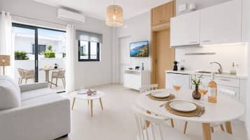 Uma sala de estar moderna com um sofá, uma mesa de jantar para dois e uma área de cozinha com armários brancos e uma televisão.