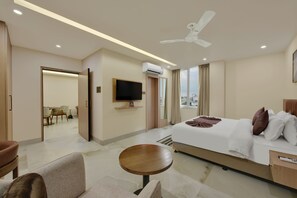 Quarto de hotel com cama, televisão, ventoinha no teto, área de refeições e vista para a cidade.