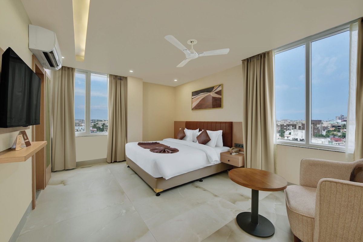 Quarto de hotel com cama, escrivaninha, cadeira, ventoinha no teto e janelas amplas com vista para a cidade.