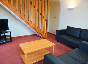 Sala de estar com escada de madeira, sofá preto, mesa de centro de madeira e televisão em móvel.