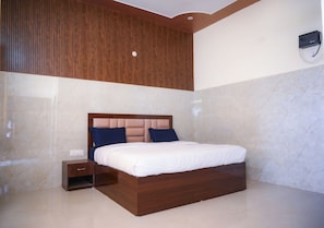 Quarto com cabeceira de madeira, cama com roupa de cama branca, mesa de cabeceira e carregador fixado na parede.