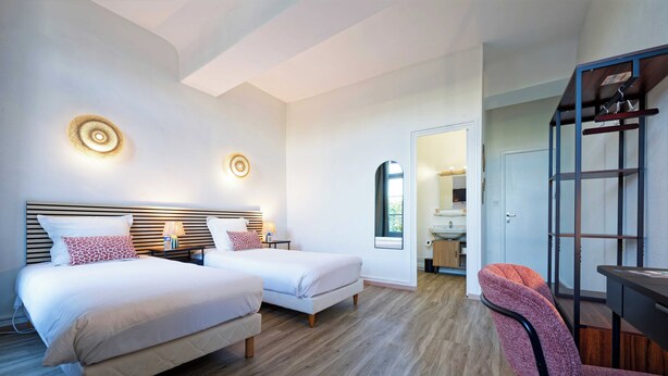 Quarto de hotel com duas camas, uma mesa, uma cadeira e um banheiro visível por uma porta aberta.
