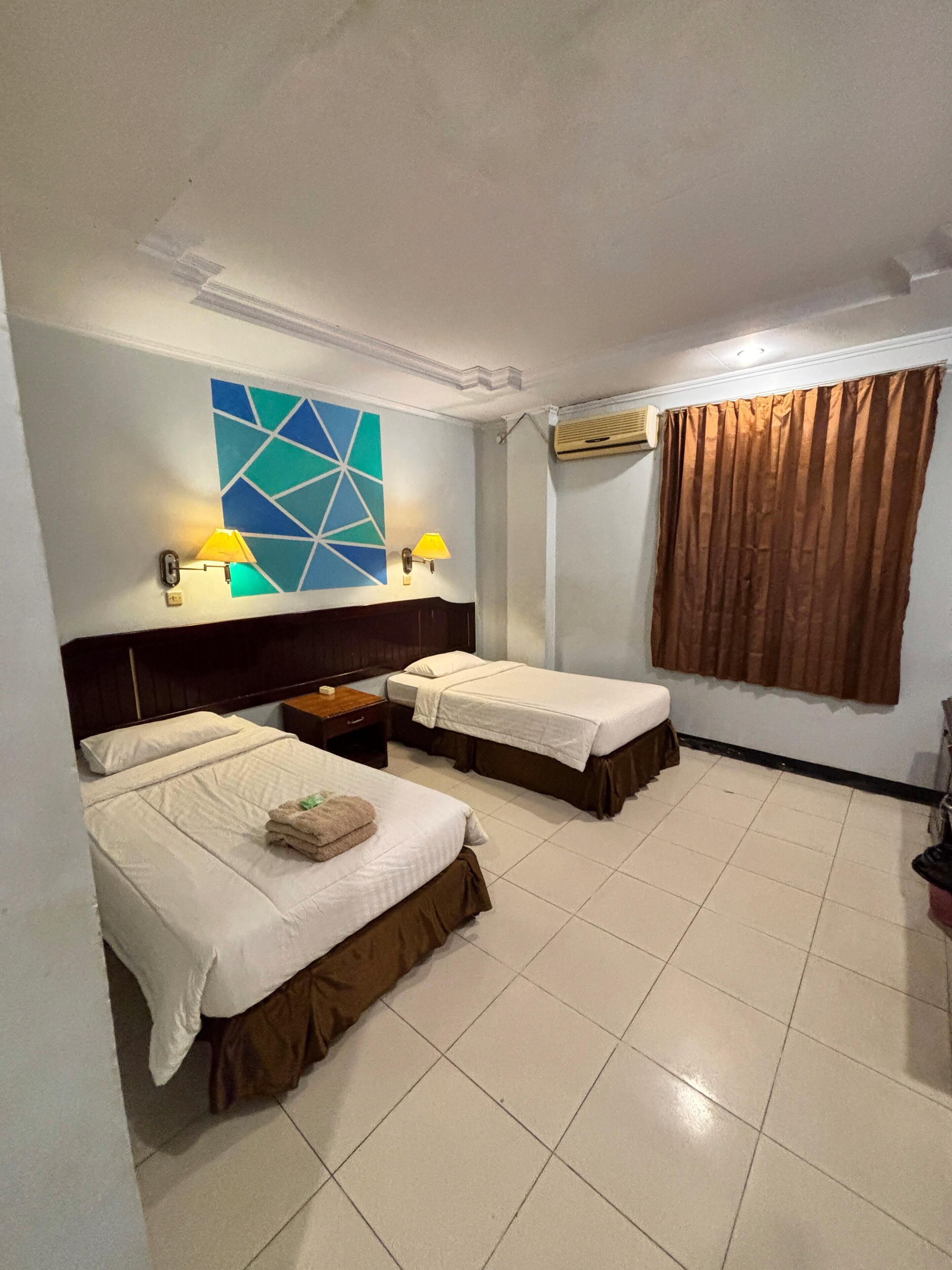 Quarto de hotel com duas camas, uma obra de arte geométrica na parede, uma janela com cortinas e um ar condicionado fixado na parede.