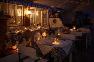 Uma área de refeições ao ar livre com mesas preparadas para uma refeição, iluminada por velas e lanternas, sob uma estrutura coberta com janelas.