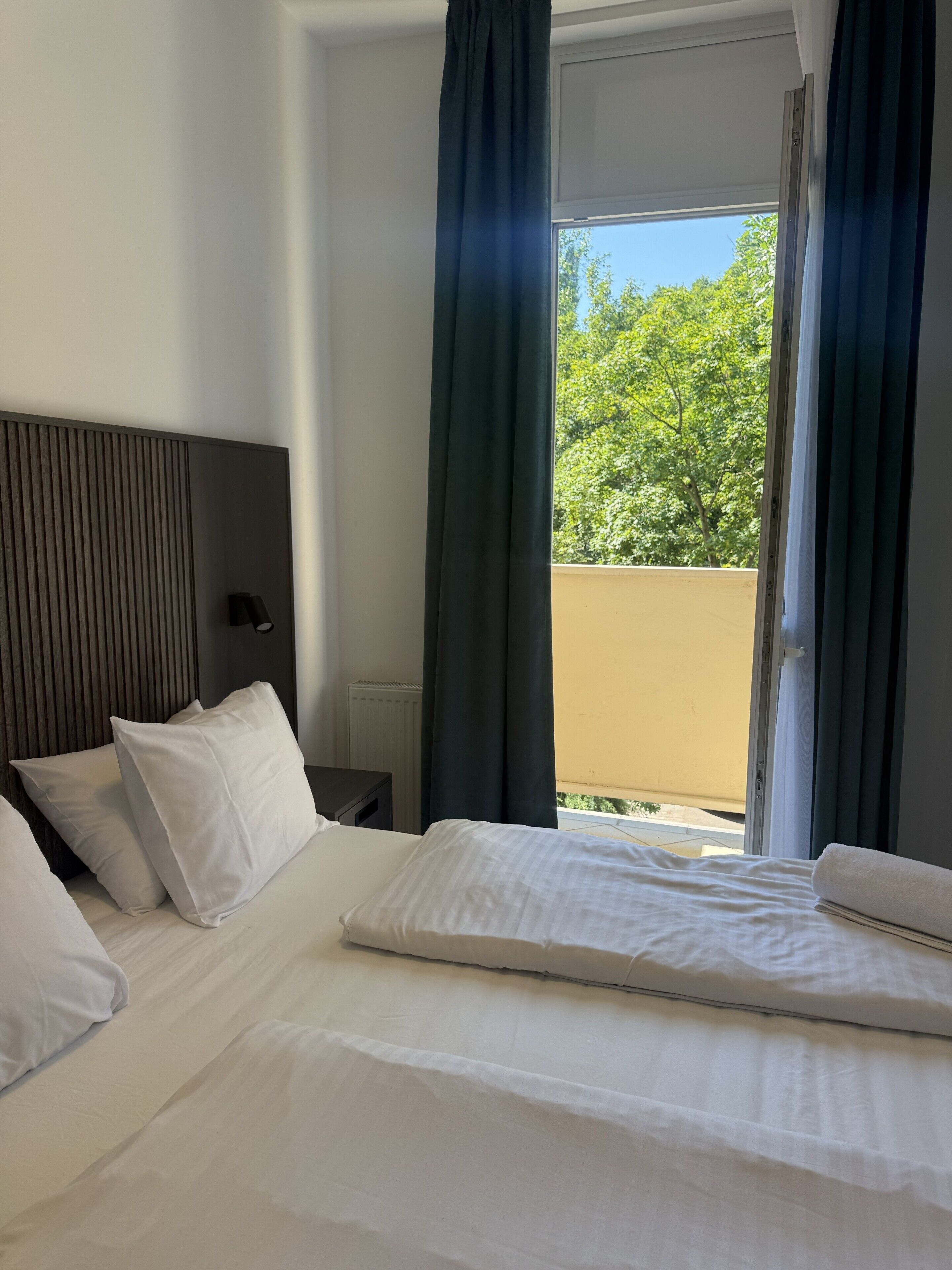 Quarto de hotel com cama, mesa de cabeceira, luminária e vista para o verde lá fora.