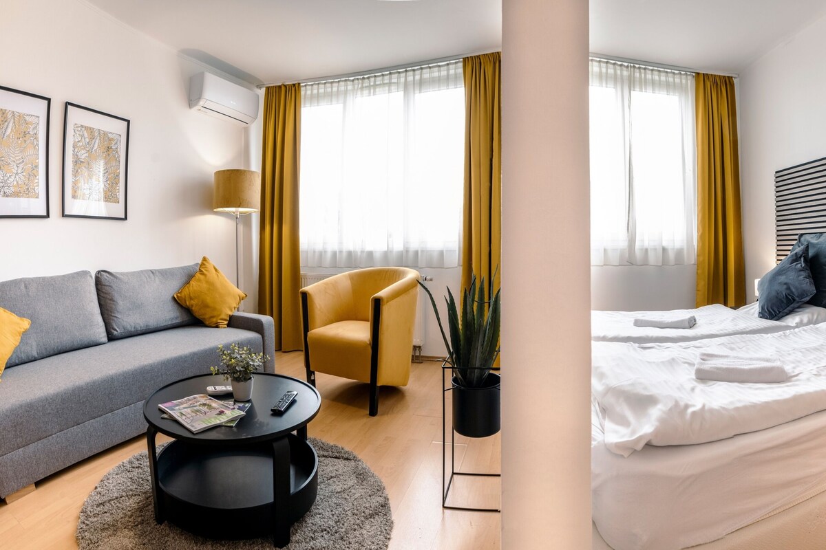 Quarto de hotel moderno com sofá, poltrona, mesa de centro redonda e uma cama com cabeceira.