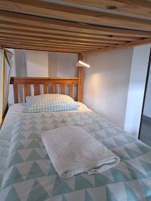 Uma cama de solteiro com colcha estampada em padrão geométrico, cabeceira de madeira e uma toalha branca sobre a cama.