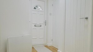 Uma porta branca com uma pequena janela e uma abertura para correspondência, dando acesso a um quarto com piso de madeira.