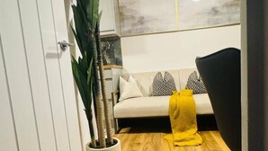 Quarto com piso de madeira, porta branca, planta em vaso, sofá com almofadas e uma cadeira.