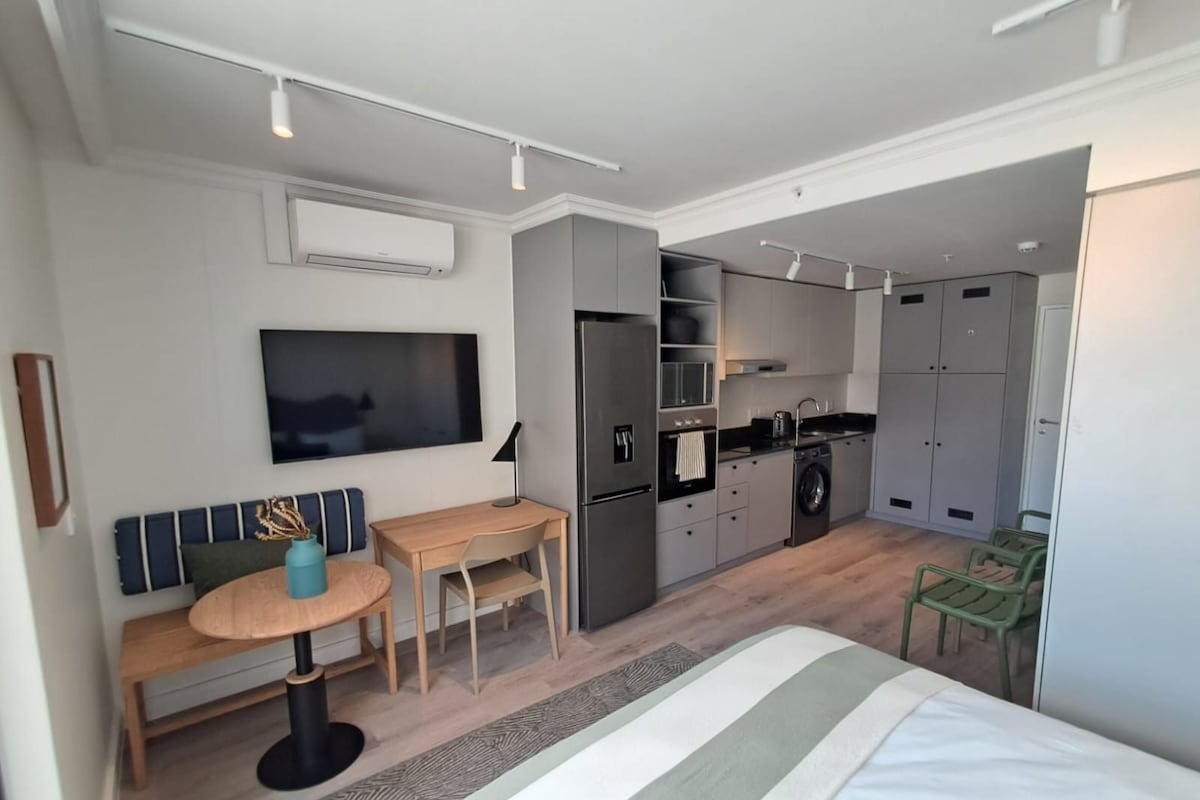 Quarto de hotel moderno com cama, área de refeições, cozinha compacta e TV.